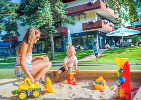 Uma área de lazer com areia, brinquedos infantis e um edifício com varandas ao fundo.