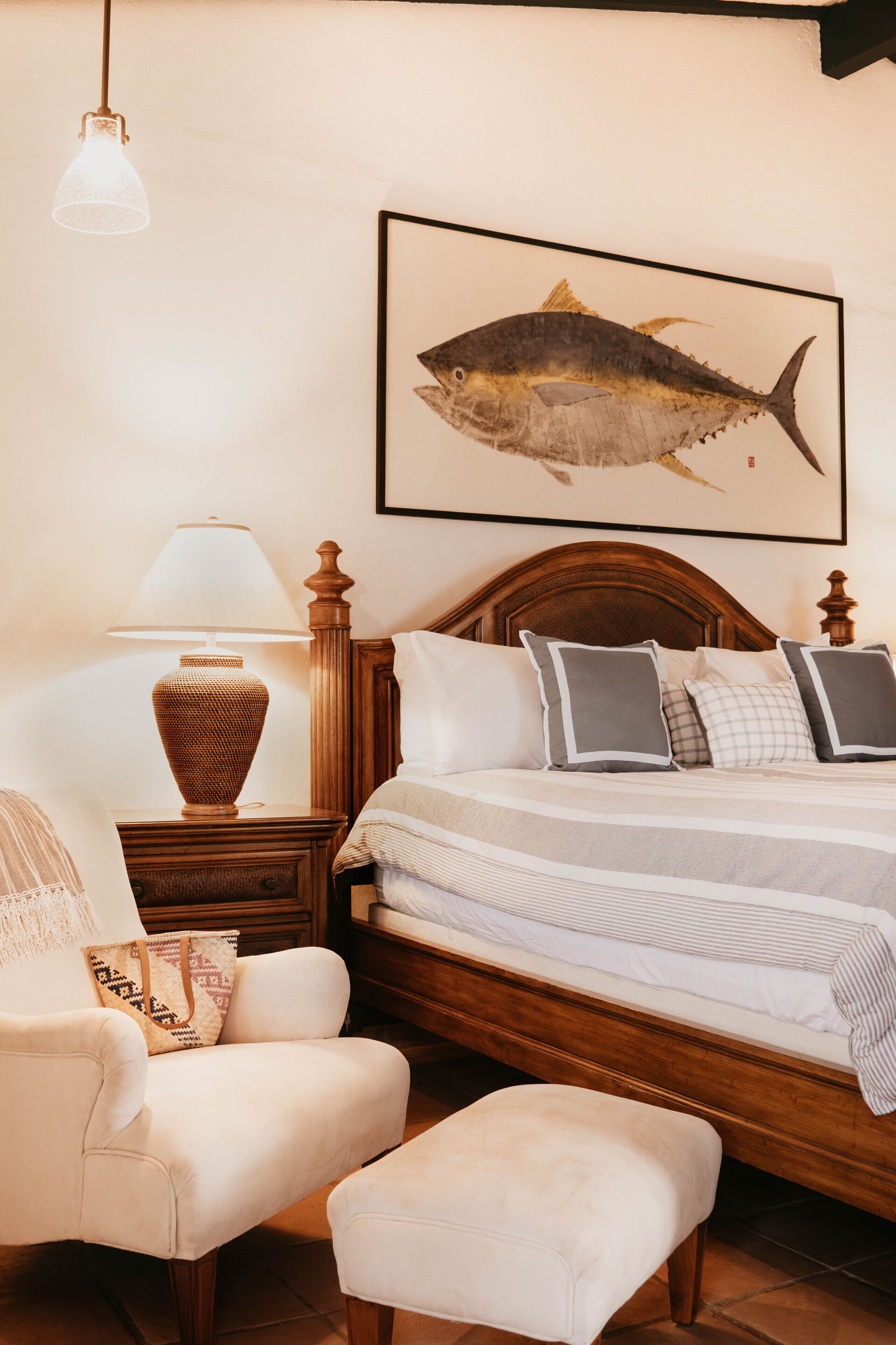 Quarto com cama de madeira, mesa de cabeceira com abajur, uma cadeira e um quadro de peixe na parede.