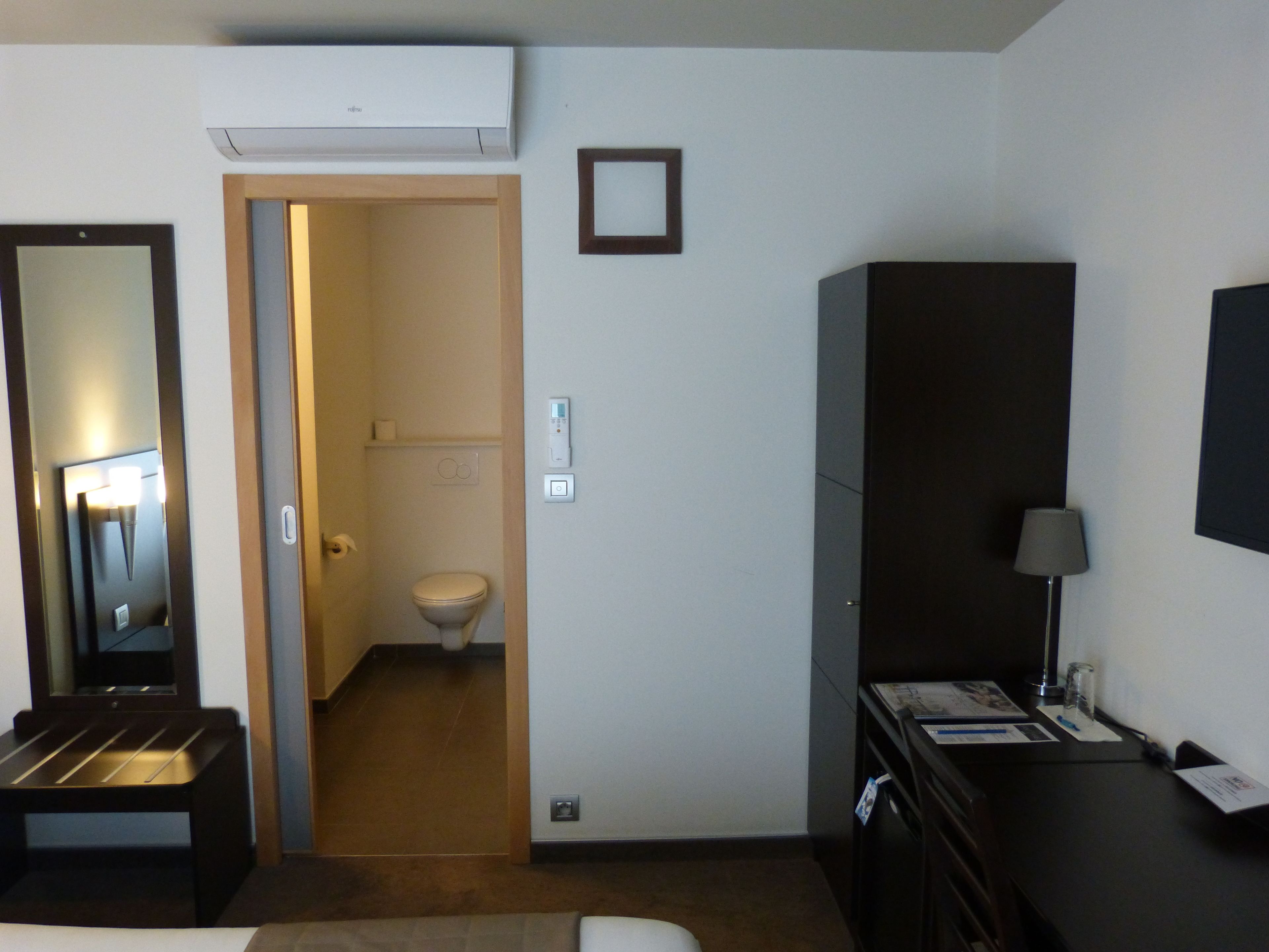 Quarto de hotel com cama, escrivaninha, espelho, guarda-roupa, sanita e área de duche.