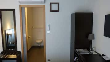 Quarto de hotel com cama, escrivaninha, espelho, guarda-roupa, sanita e área de duche.