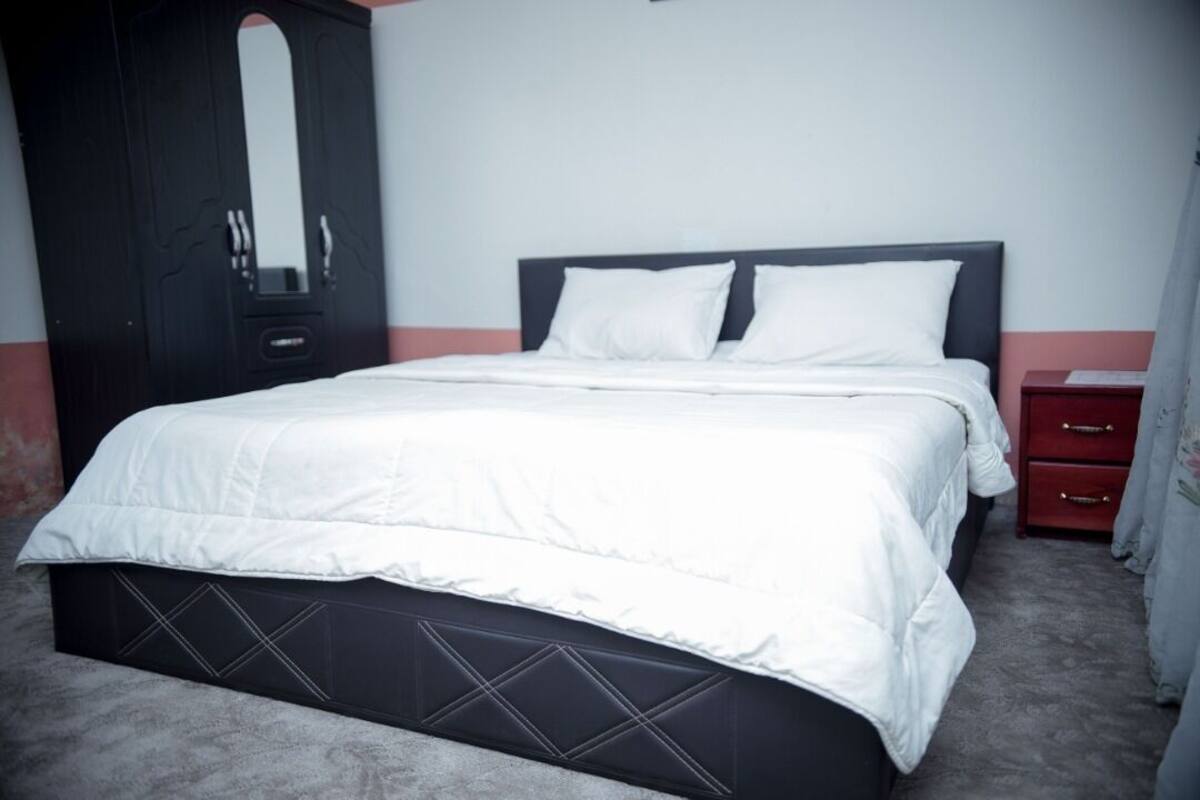 Cama bem arrumada com cabeceira escura e roupa de cama branca, mesa de cabeceira vermelha com gavetas, e guarda-roupa preto com espelho.
