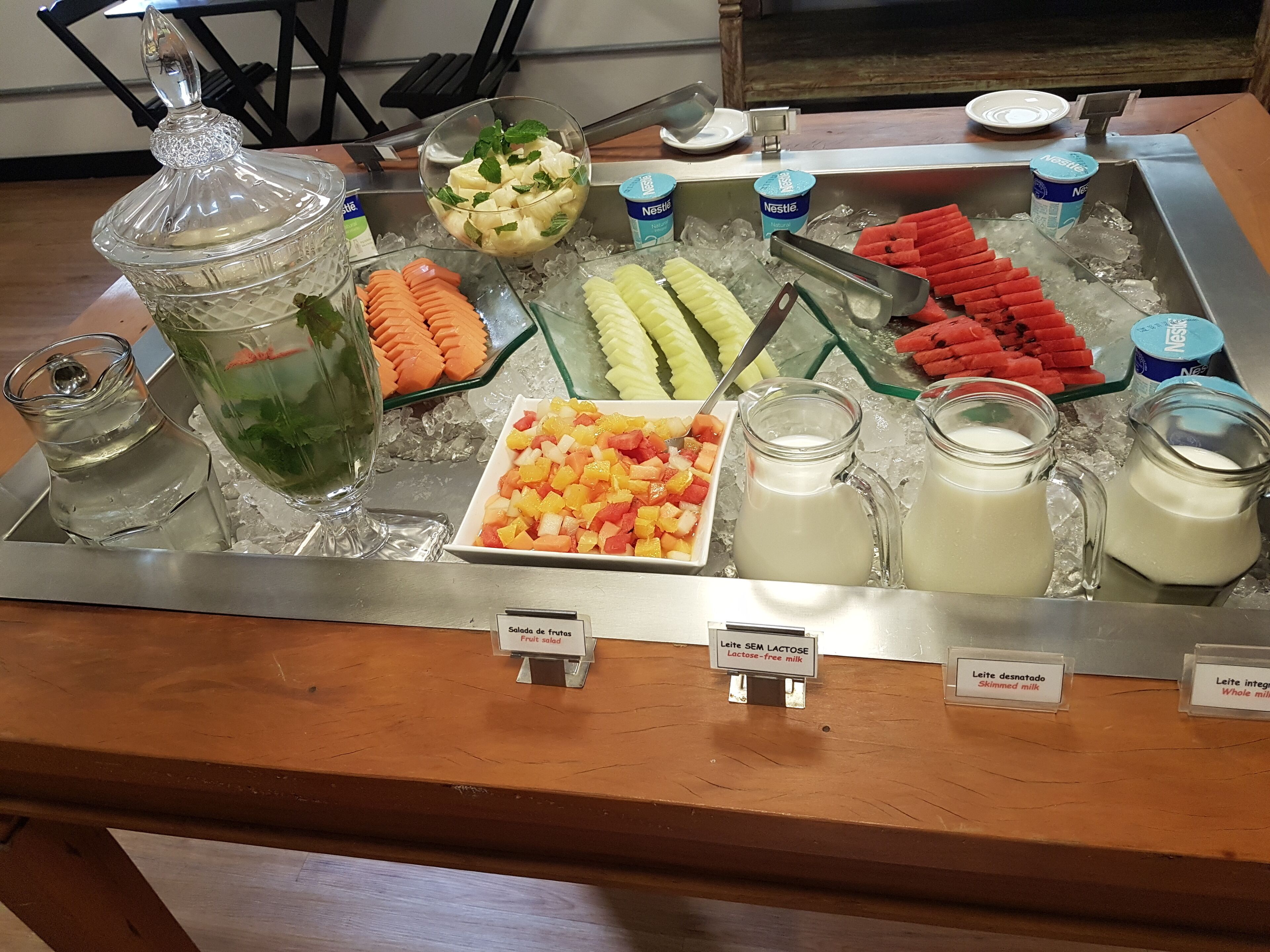 Buffet com diversos alimentos e bebidas, incluindo salada de frutas, frutas fatiadas e diferentes tipos de leite.