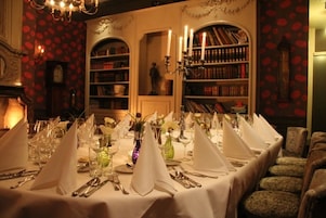 Uma sala de jantar formal com uma longa mesa posta para refeição, cercada por estantes de livros e um relógio.