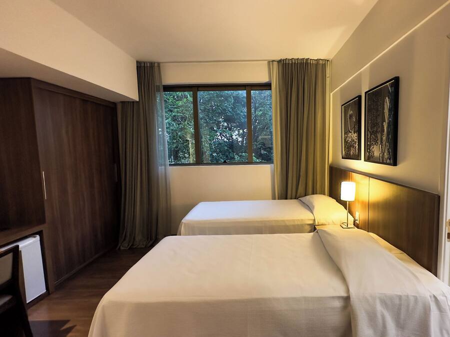 Quarto de hotel com duas camas, uma escrivaninha, um guarda-roupa e uma janela com cortinas.