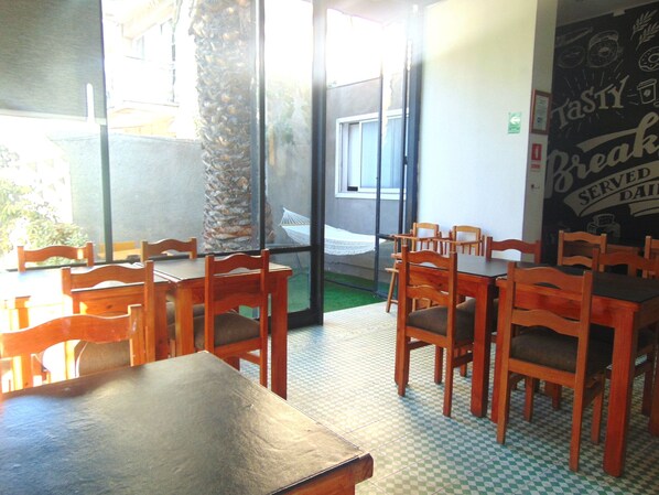 Restaurante com mesas e cadeiras de madeira, chão xadrez e uma grande janela com vista para o exterior.