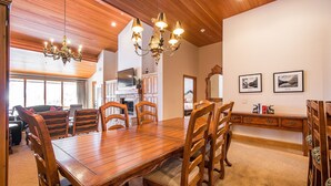 Uma sala de jantar com mesa e cadeiras de madeira, um candelabro e uma cozinha ao fundo.