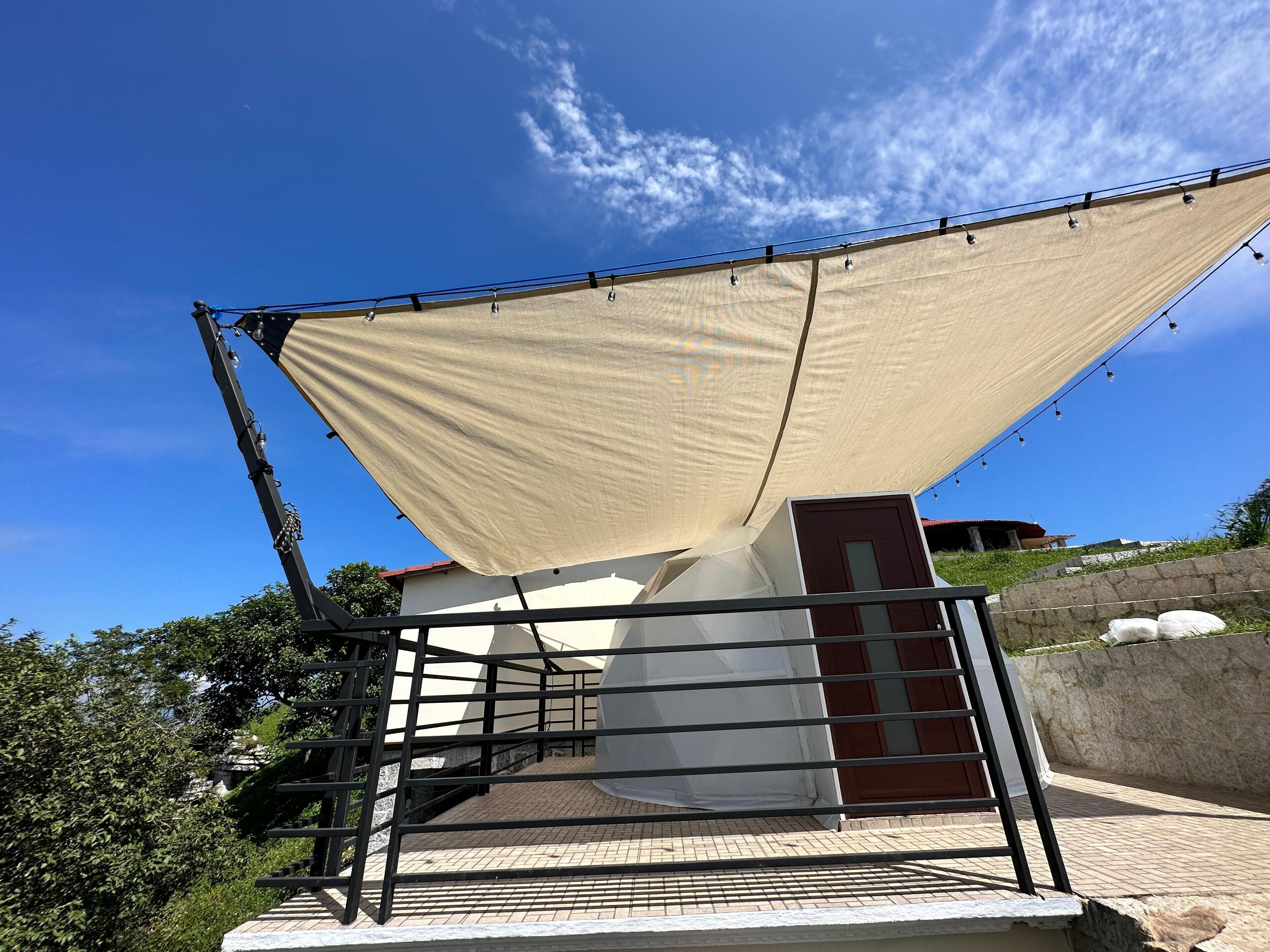 Uma área externa coberta com uma estrutura de tenda branca, corrimão de metal e um muro de pedra ao fundo.