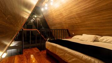 Um quarto com teto inclinado de madeira, uma cama com roupa de cama branca, uma escrivaninha de madeira e uma escada.