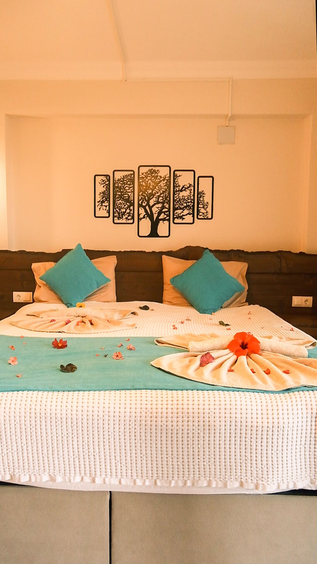 Uma cama bem arrumada com colcha verde-água, dois travesseiros azuis e uma toalha decorativa com uma flor vermelha.