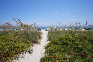 Um caminho de areia na praia, com guarda-sóis e pessoas ao longe.