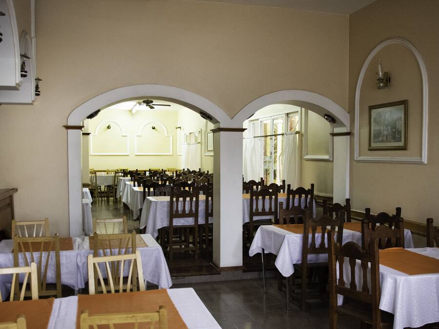 Uma sala de jantar com várias mesas e cadeiras, portas em arco e vista para outro ambiente.