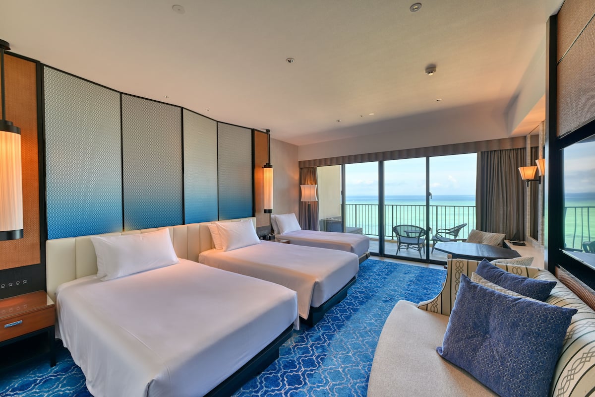 Quarto de hotel com duas camas, um sofá, uma varanda com mesa e cadeiras e vista para o mar.