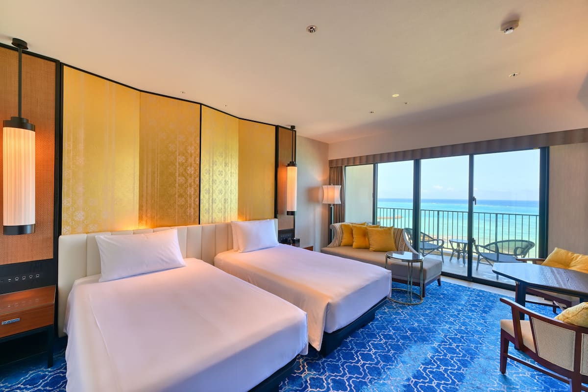 Quarto de hotel com duas camas, varanda com vista para o mar e área de refeições com almofadas amarelas.