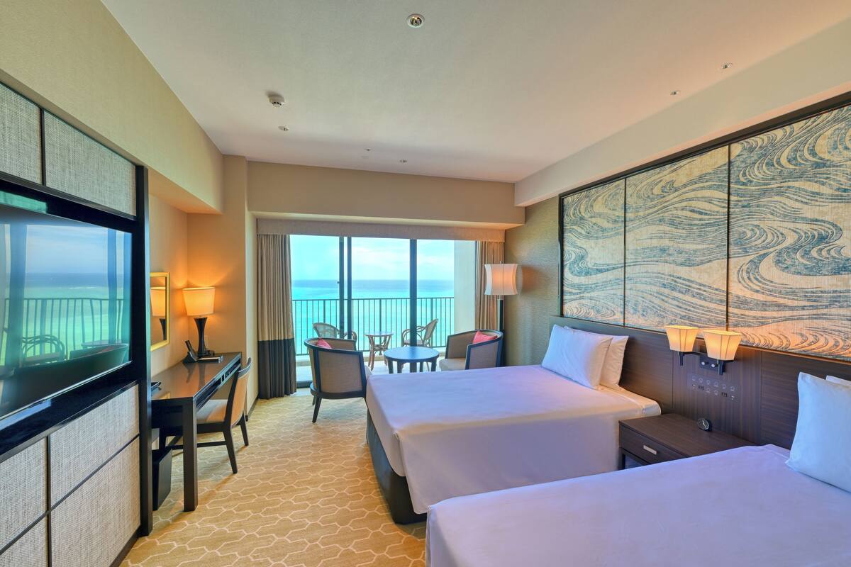 Quarto de hotel com duas camas, uma escrivaninha, cadeiras, televisão e uma janela grande com vista para o mar.
