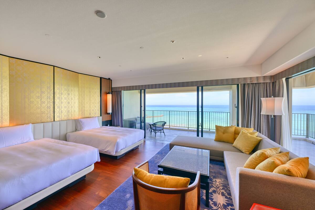 Quarto de hotel com duas camas, um sofá, uma mesa de jantar e varanda com vista para o mar.