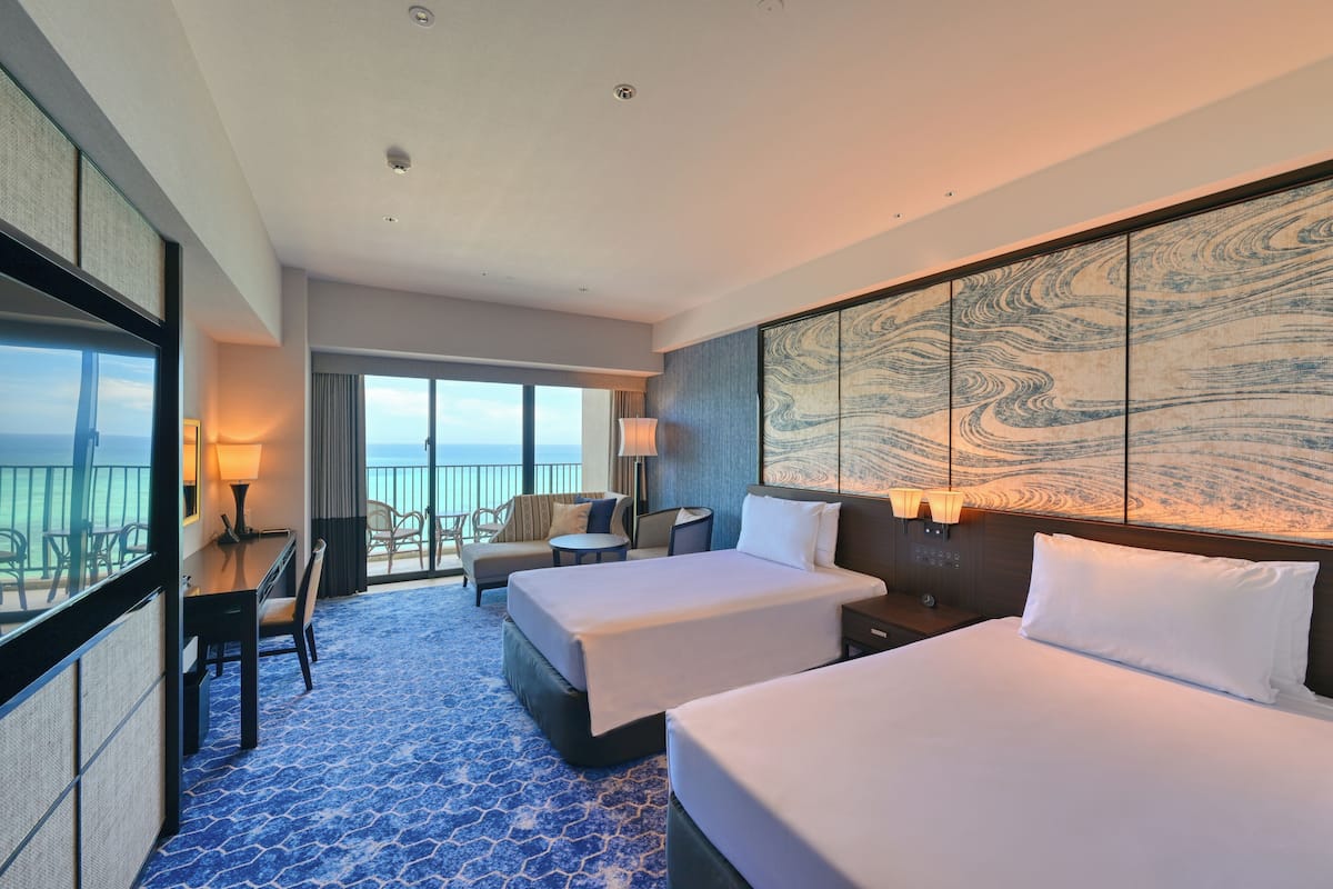 Quarto de hotel com duas camas, uma escrivaninha, uma cadeira, uma mesinha e uma janela grande com vista para o mar.