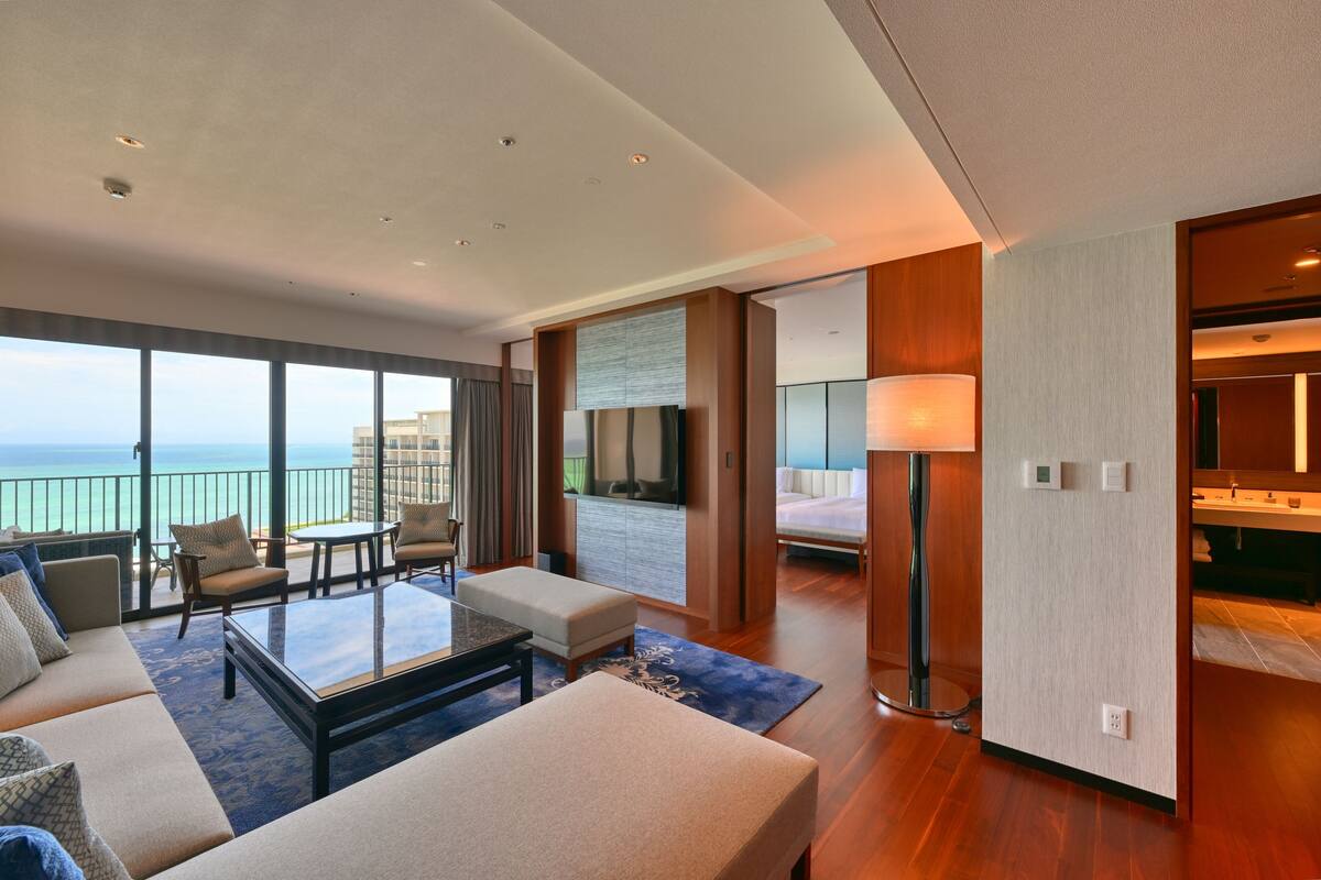 Quarto de hotel moderno com varanda, sofá, mesa de centro, televisão e banheiro com espelho e pia.