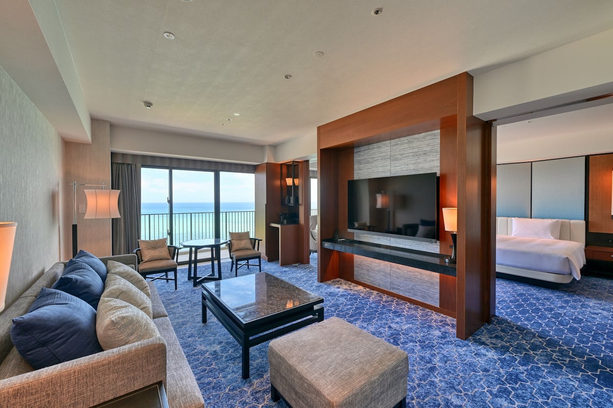 Quarto de hotel com uma televisão grande, um sofá, uma mesa de centro e uma varanda com vista para o oceano.