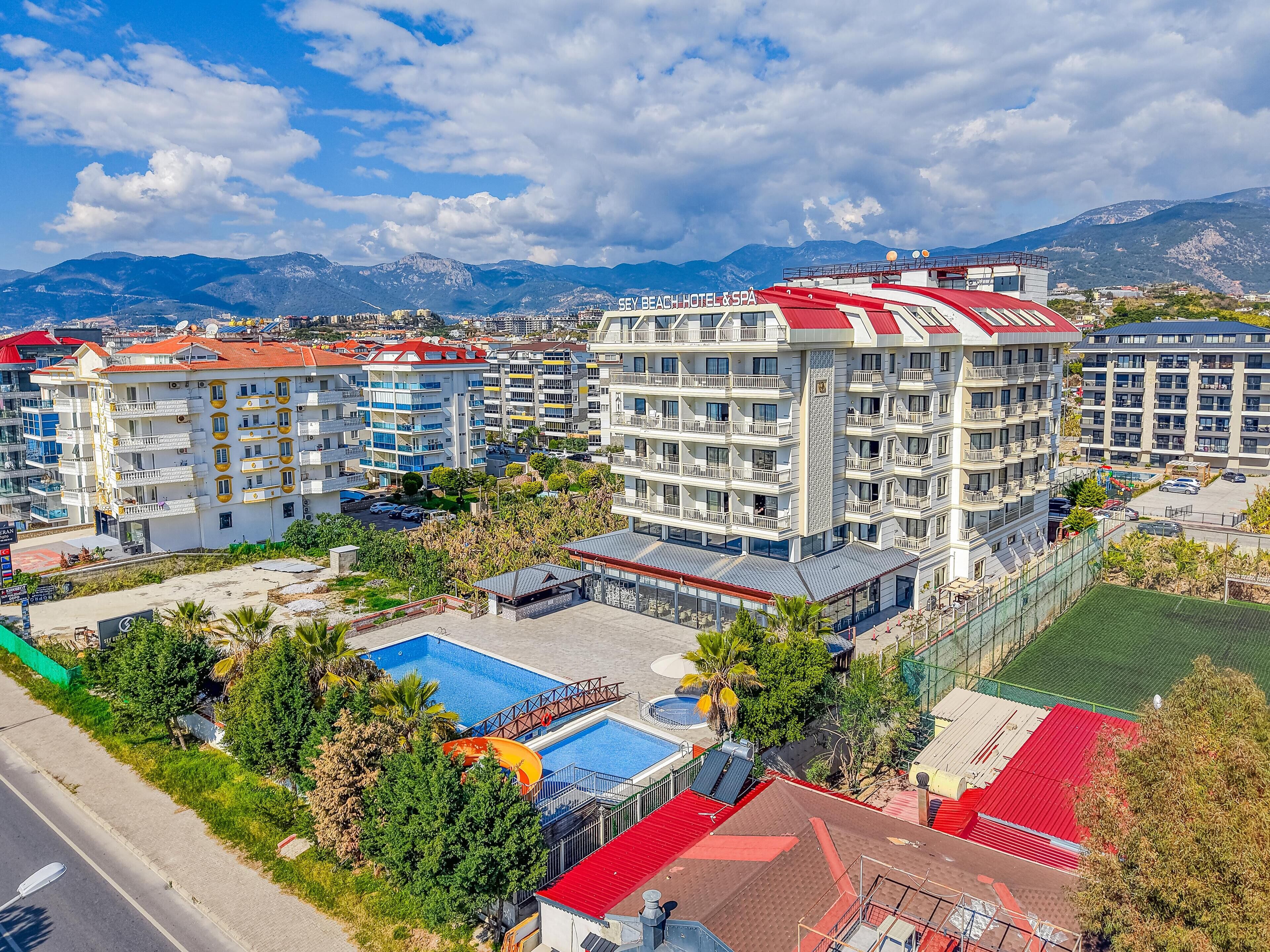 Um hotel com vários andares, varandas, piscina e um edifício com telhado vermelho.