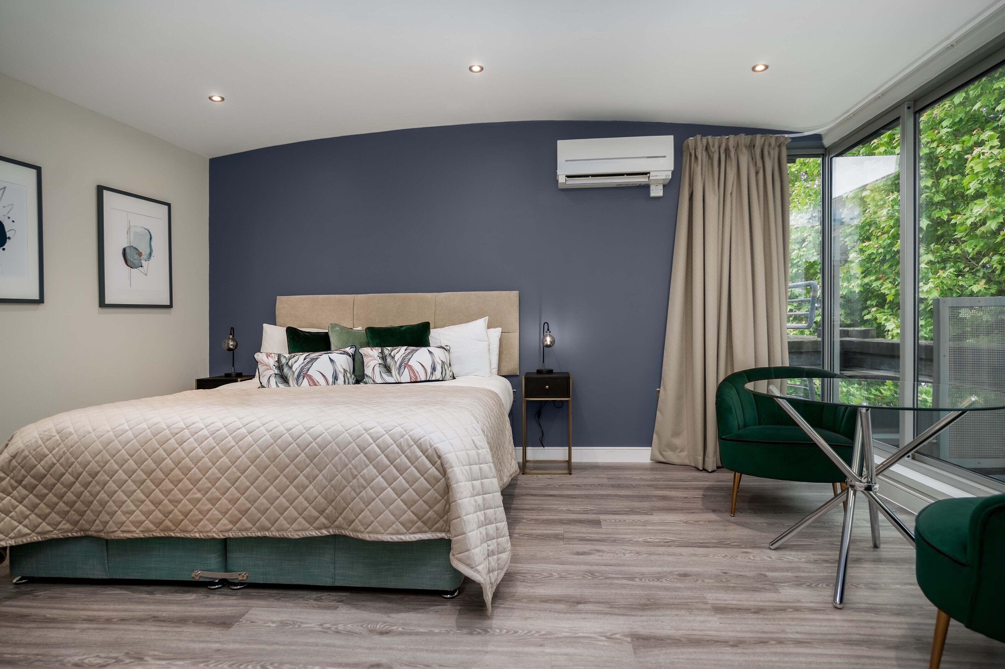 Quarto moderno com cama grande, mesa de cabeceira, cadeira verde e mesa de vidro.