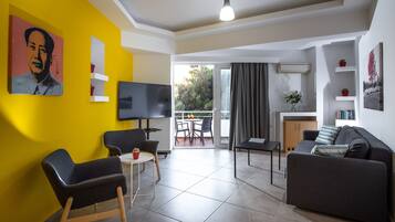Uma sala de estar moderna com uma parede de destaque amarela, uma TV de tela plana, um sofá, uma mesa de centro e uma área de jantar com vista para o verde lá fora.