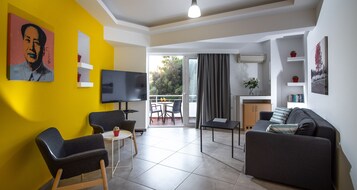 Uma sala de estar moderna com uma parede de destaque amarela, uma TV de tela plana, um sofá, uma mesa de centro e uma área de jantar com vista para o verde lá fora.