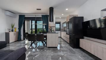 Uma cozinha moderna com uma televisão grande, um espaço de refeições com mesa e cadeiras, e vista para o exterior através de amplas janelas.