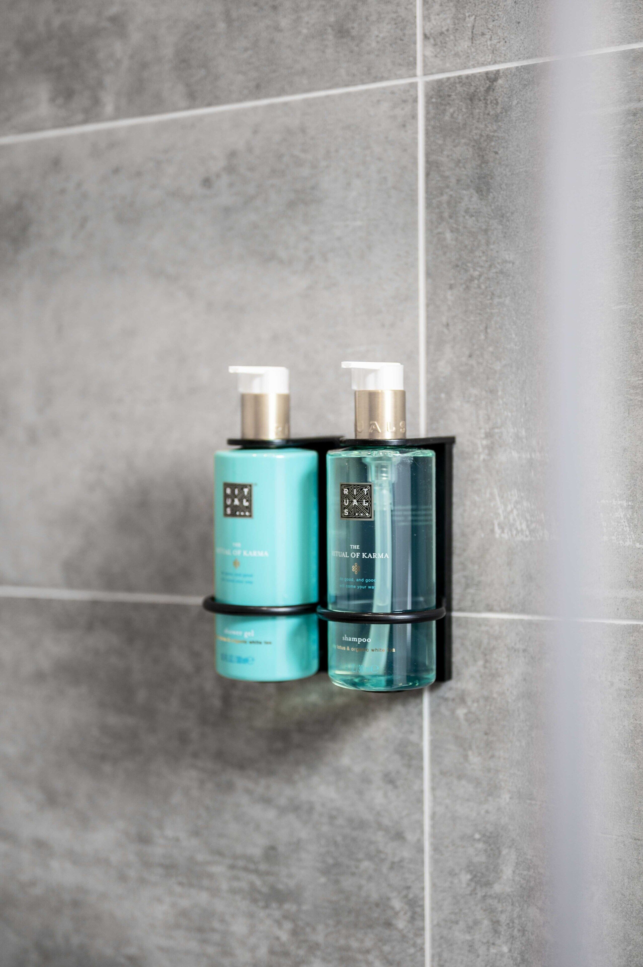Duas embalagens de shampoo e condicionador The Ritual of Karma fixadas numa parede revestida a azulejos.
