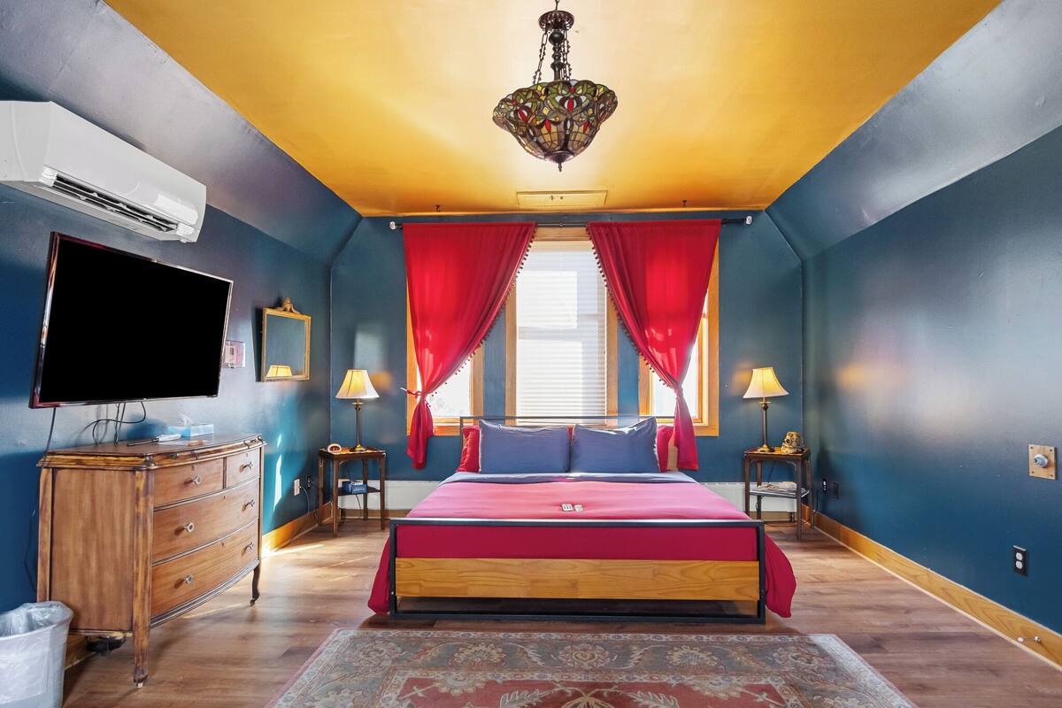 Quarto com cama, cortinas, lustre, televisão e cômoda.
