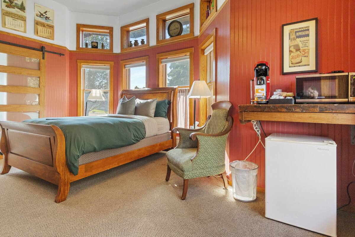 Quarto com cama de madeira, poltrona verde, frigorífico branco pequeno e secretária de madeira com candeeiro e relógio.