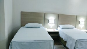 Quarto de hotel com duas camas de solteiro, cada uma com cabeceira e mesa de cabeceira. Duas luminárias fixadas na parede em cada mesa.