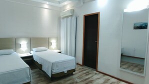 Quarto de hotel com duas camas de solteiro, mesa de cabeceira com abajur, ar condicionado, porta com espelho e uma pequena imagem na parede.