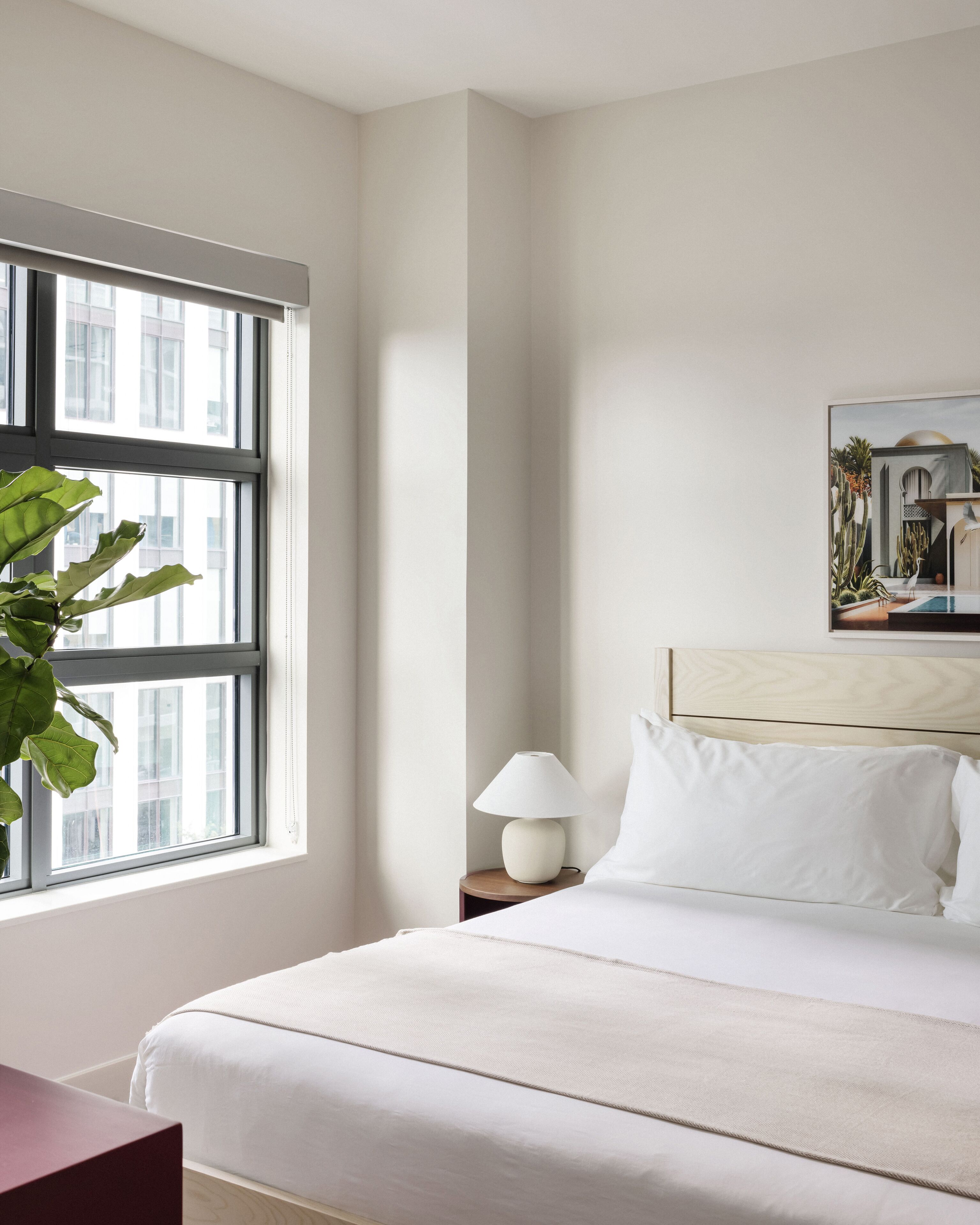 Quarto de hotel com uma janela grande, uma cama com roupa de cama branca, uma mesinha de cabeceira com abajur e um quadro na parede.