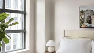 Quarto de hotel com uma janela grande, uma cama com roupa de cama branca, uma mesinha de cabeceira com abajur e um quadro na parede.