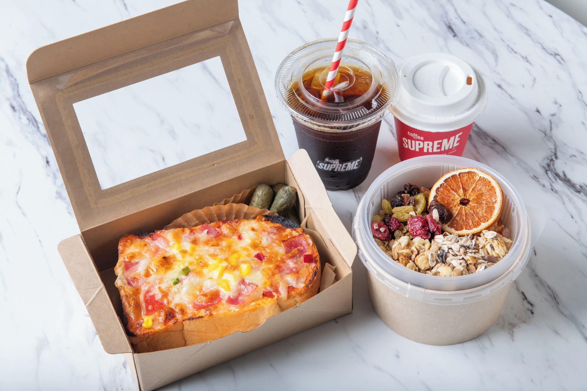 Uma caixa de cartão com uma fatia de pizza, uma chávena de café gelado e um recipiente de cereais com fruta e frutos secos.
