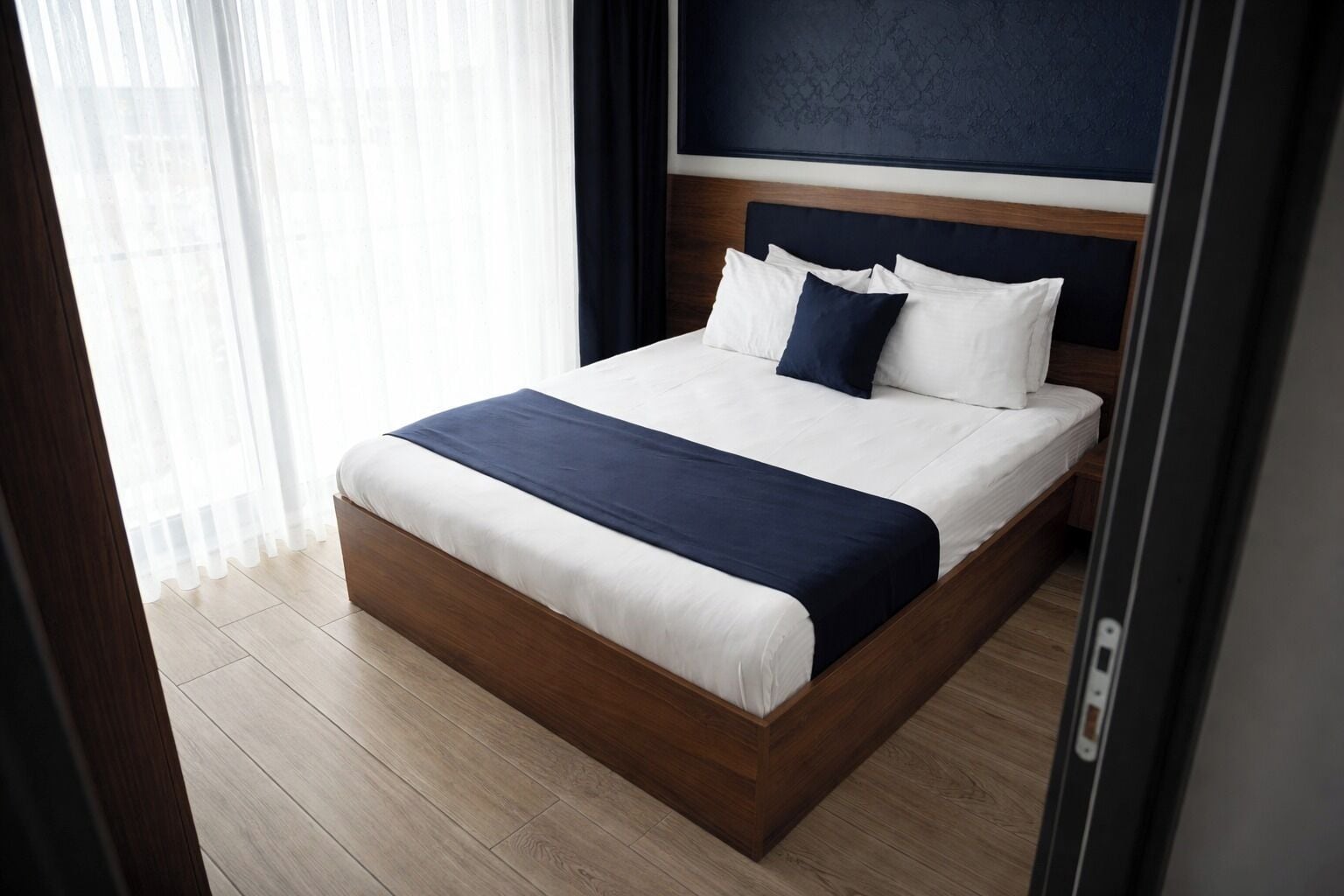 Quarto de hotel com estrutura de cama em madeira, roupa de cama branca e cabeceira escura.