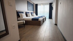 Quarto de hotel com piso de madeira, uma cama com cabeceira, mesa de cabeceira e uma janela com cortinas.