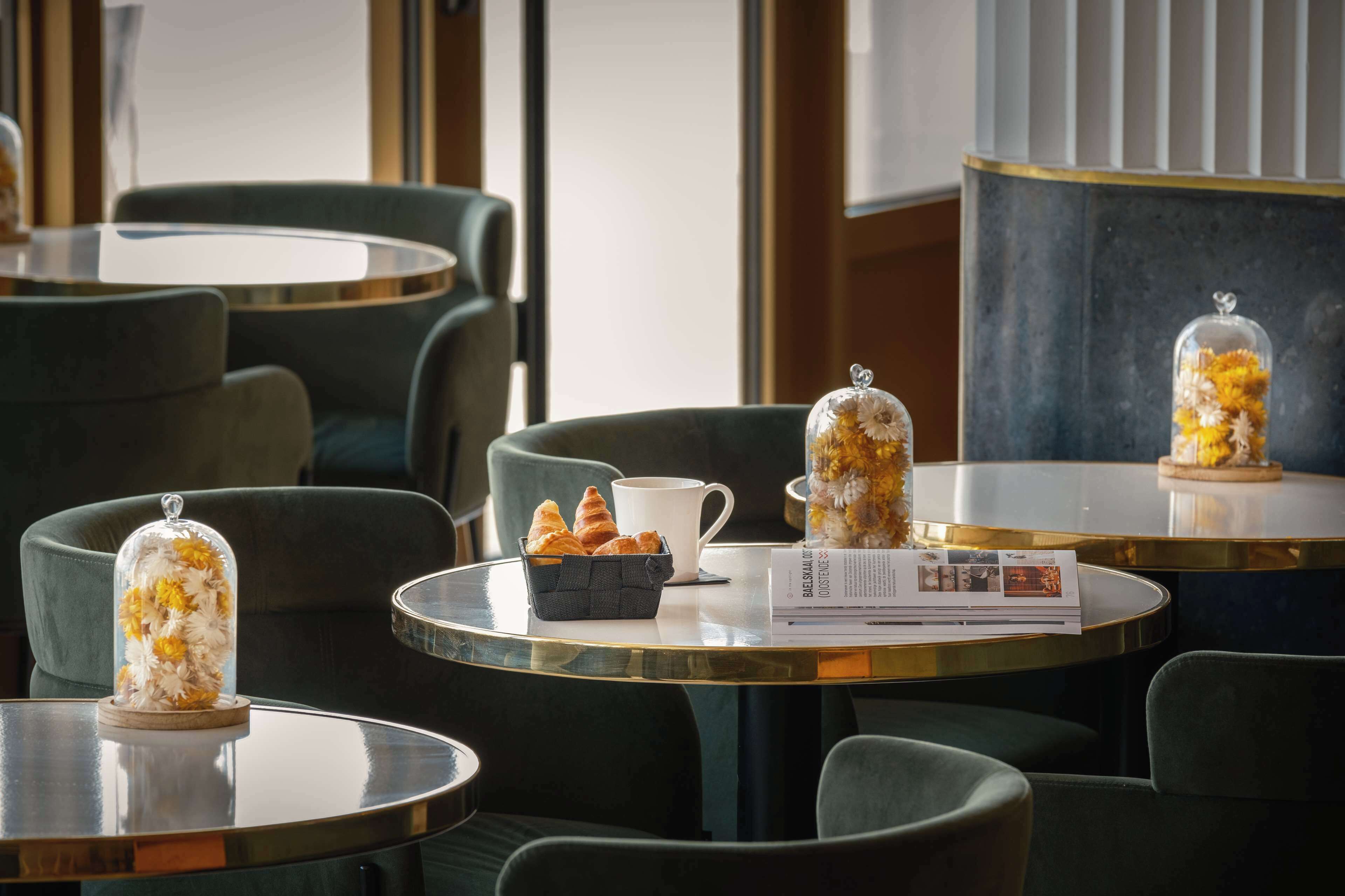 Um ambiente acolhedor de café com mesas redondas, cadeiras estofadas em verde, uma mesa de centro de vidro, uma chávena branca, uma cesta de croissants e um menu.