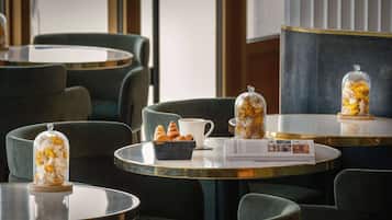 Um ambiente acolhedor de café com mesas redondas, cadeiras estofadas em verde, uma mesa de centro de vidro, uma chávena branca, uma cesta de croissants e um menu.
