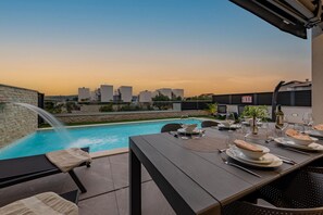 Uma área de refeições no terraço com piscina, mesas ao ar livre e vista para a cidade.