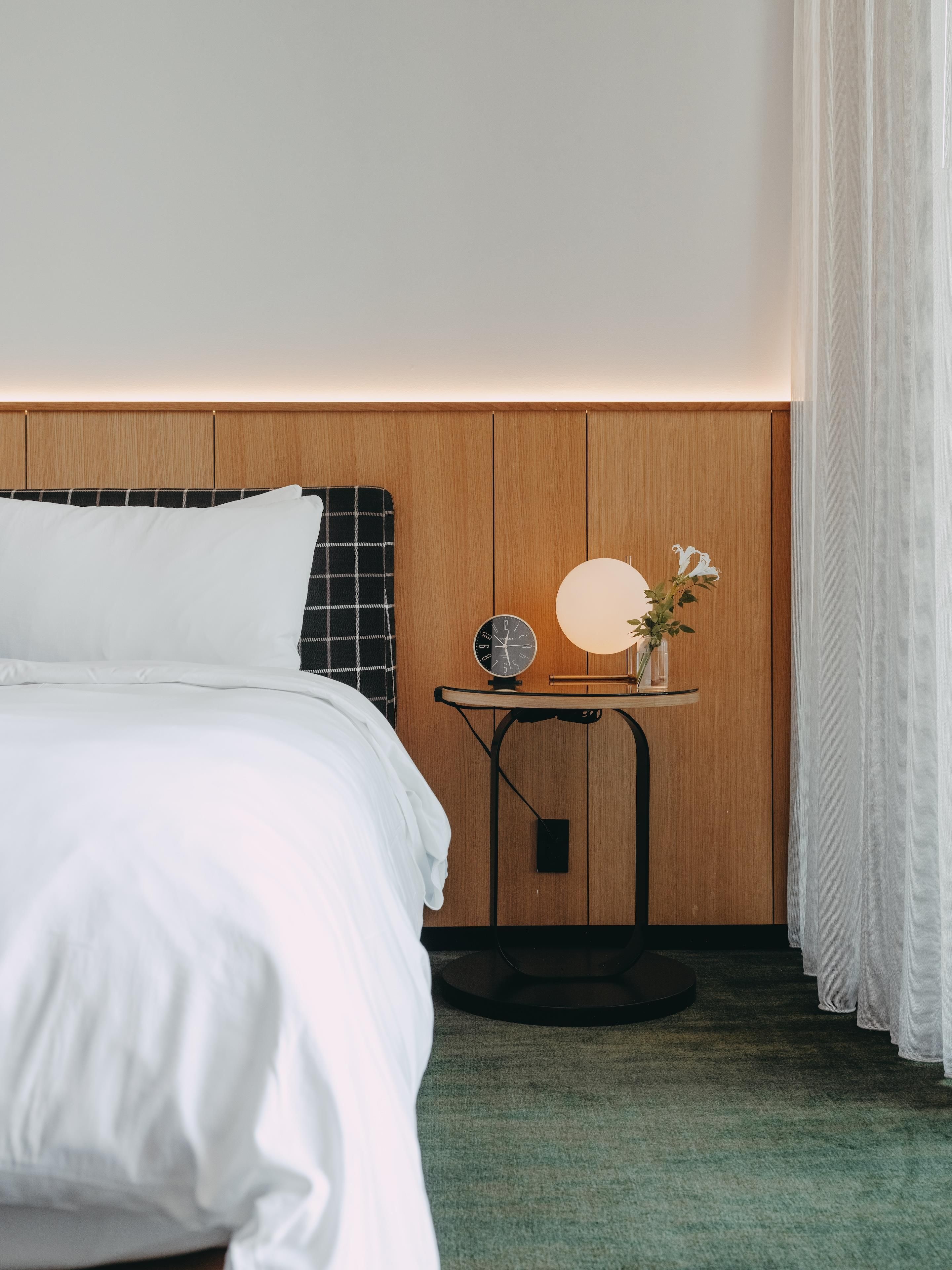 Cama bem arrumada com roupa de cama branca, mesa de cabeceira com relógio e um vaso de flores, e cabeceira de madeira.
