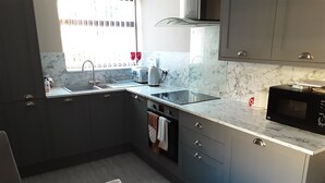 Uma cozinha compacta com armários escuros, bancada de mármore, pia, micro-ondas e janela com persianas.