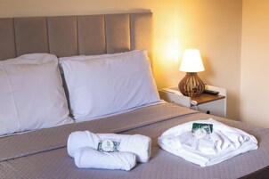Uma cama bem arrumada com roupa de cama branca, uma luminária de cabeceira e duas toalhas sobre a cama.