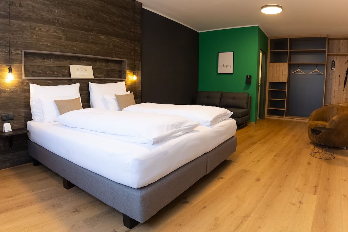 Quarto moderno com uma cama grande, um sofá cinza, um guarda-roupa de madeira e uma parede verde de destaque.