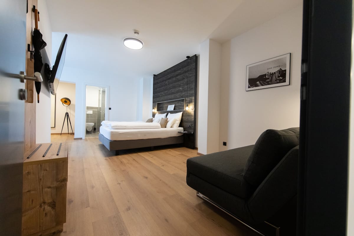 Quarto moderno com cama grande, sofá preto e piso de madeira.