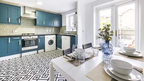 Uma cozinha moderna com armários verde-água, piso estampado e uma área de jantar para quatro pessoas, decorada com um vaso de flores.