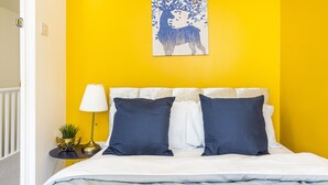 Uma cama com roupa de cama branca e azul, uma parede amarela e um quadro de um veado.