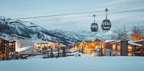 Uma cidade de montanha coberta de neve, com um teleférico, edifícios iluminados e uma mistura de arquitetura moderna e tradicional.