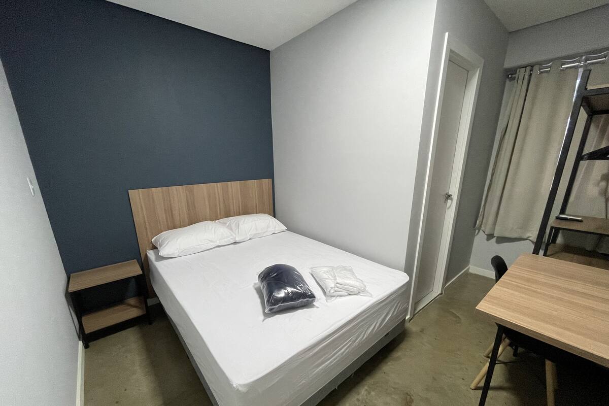 Um quarto pequeno e bem arrumado, com uma cama de solteiro, uma mesa de cabeceira de madeira, uma pequena mesa e uma porta que dá para outro cômodo.