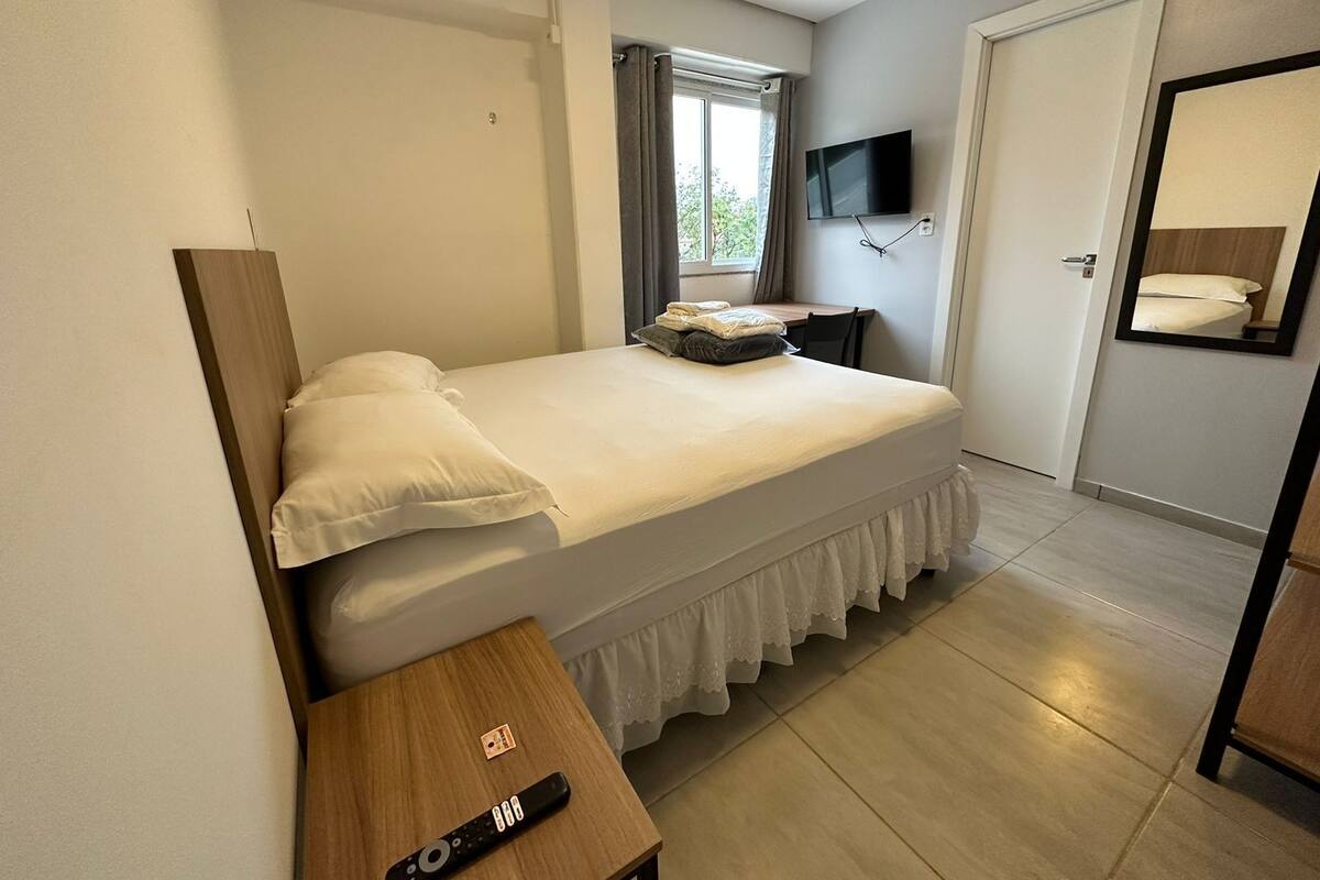 Quarto com cama, televisão presa à parede, mesa de cabeceira de madeira e espelho.